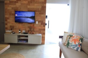 Uma sala de estar com uma televisão fixada numa parede de tijolos, um sofá com almofada florida e um radiador branco abaixo.