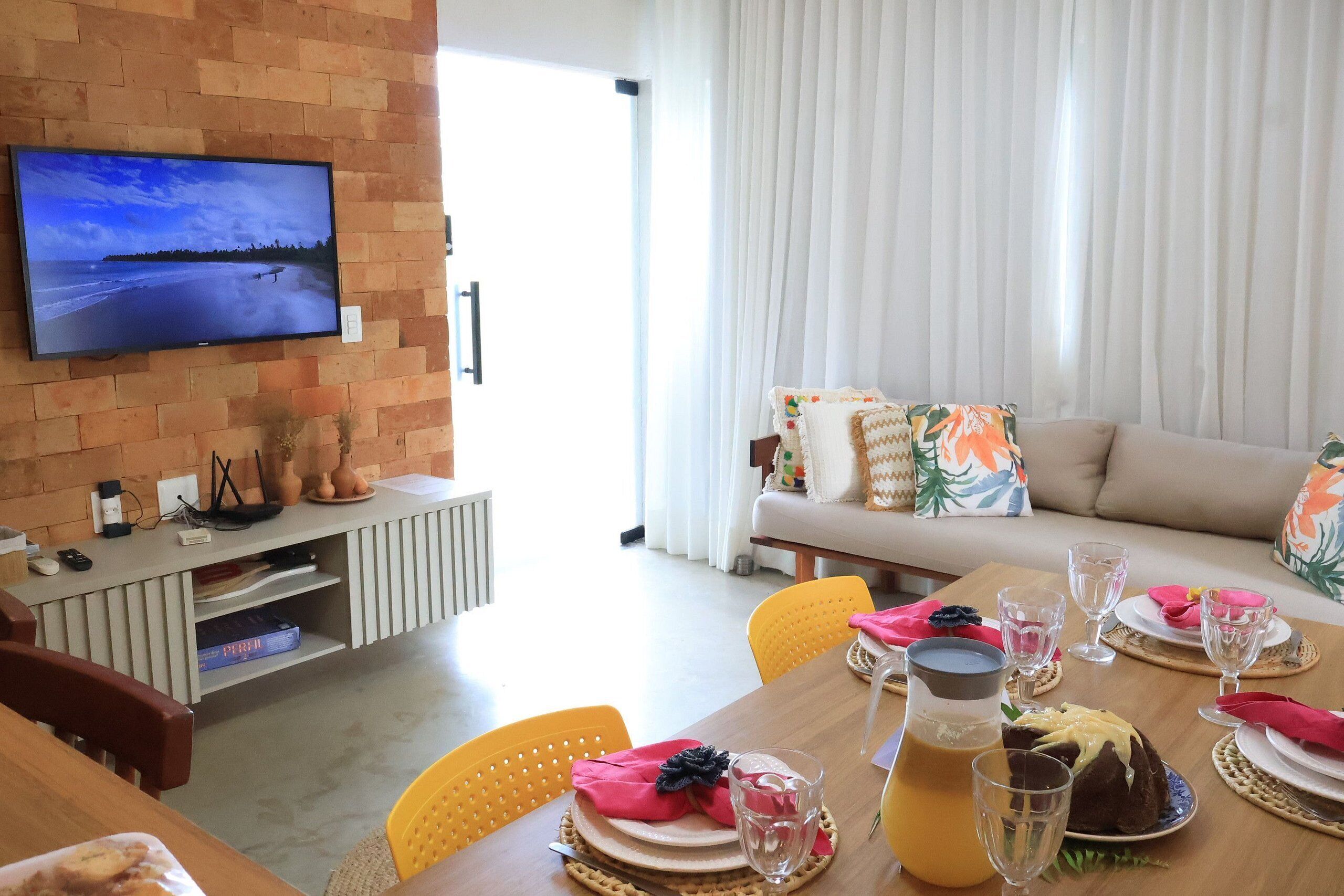 Uma sala de jantar com televisão fixada numa parede de tijolos, um sofá com almofadas estampadas e uma mesa posta para refeição.