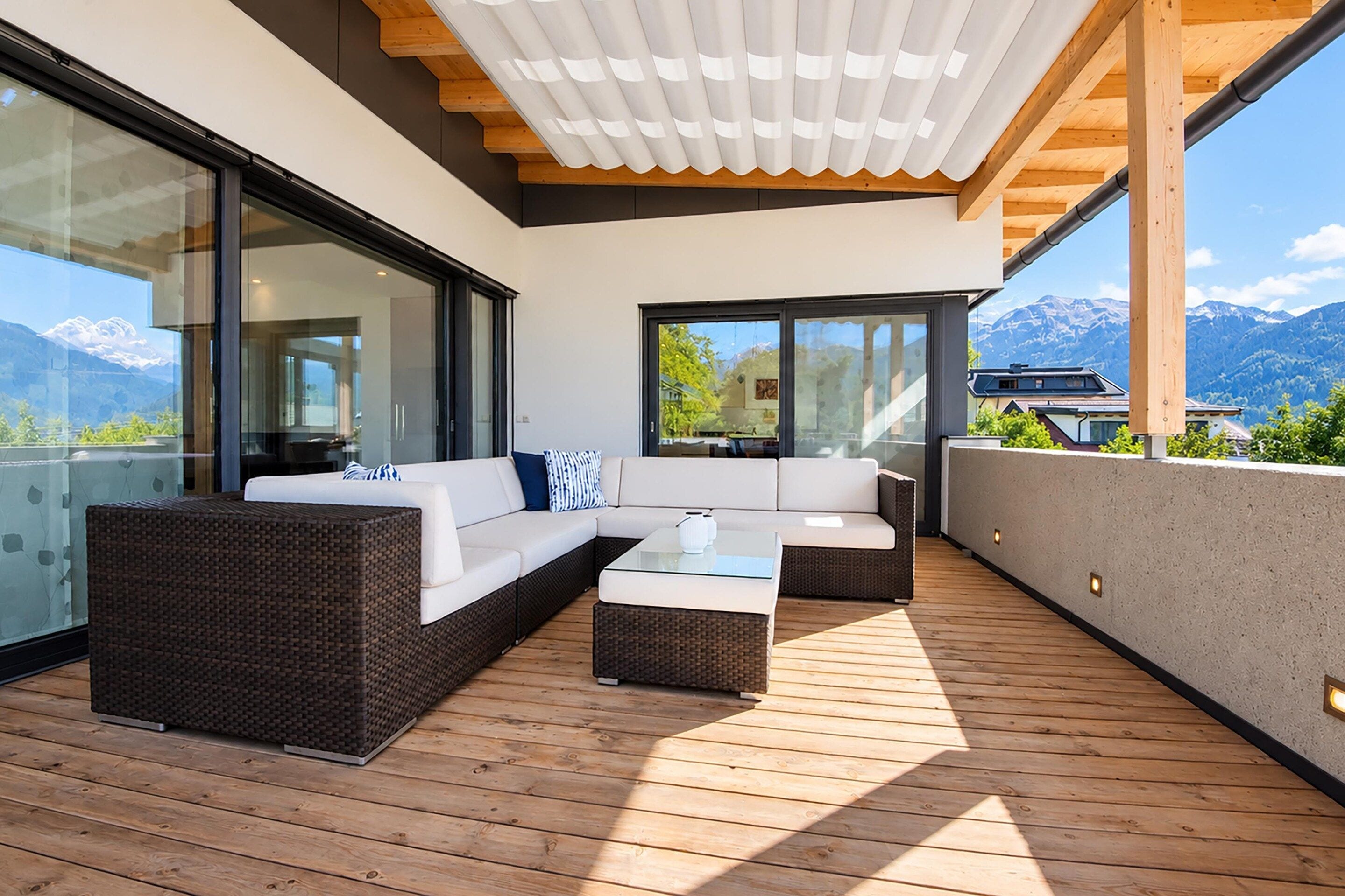 Um espaço de estar ao ar livre moderno com um deck de madeira, um sofá, uma mesa de centro e grandes janelas com vista para as montanhas.