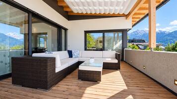 Um espaço de estar ao ar livre moderno com um deck de madeira, um sofá, uma mesa de centro e grandes janelas com vista para as montanhas.