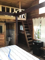 Interior de cabana com quarto em mezanino, vigas de madeira, escada para o mezanino, janela com cortinas e cama com colcha estampada.