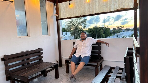 Um homem sentado num banco de madeira numa área externa coberta, com vista para a cidade.