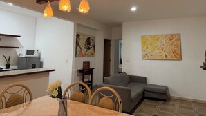 Uma sala de jantar com mesa, cadeiras, um vaso de flores, uma cozinha equipada, um sofá e obras de arte nas paredes.