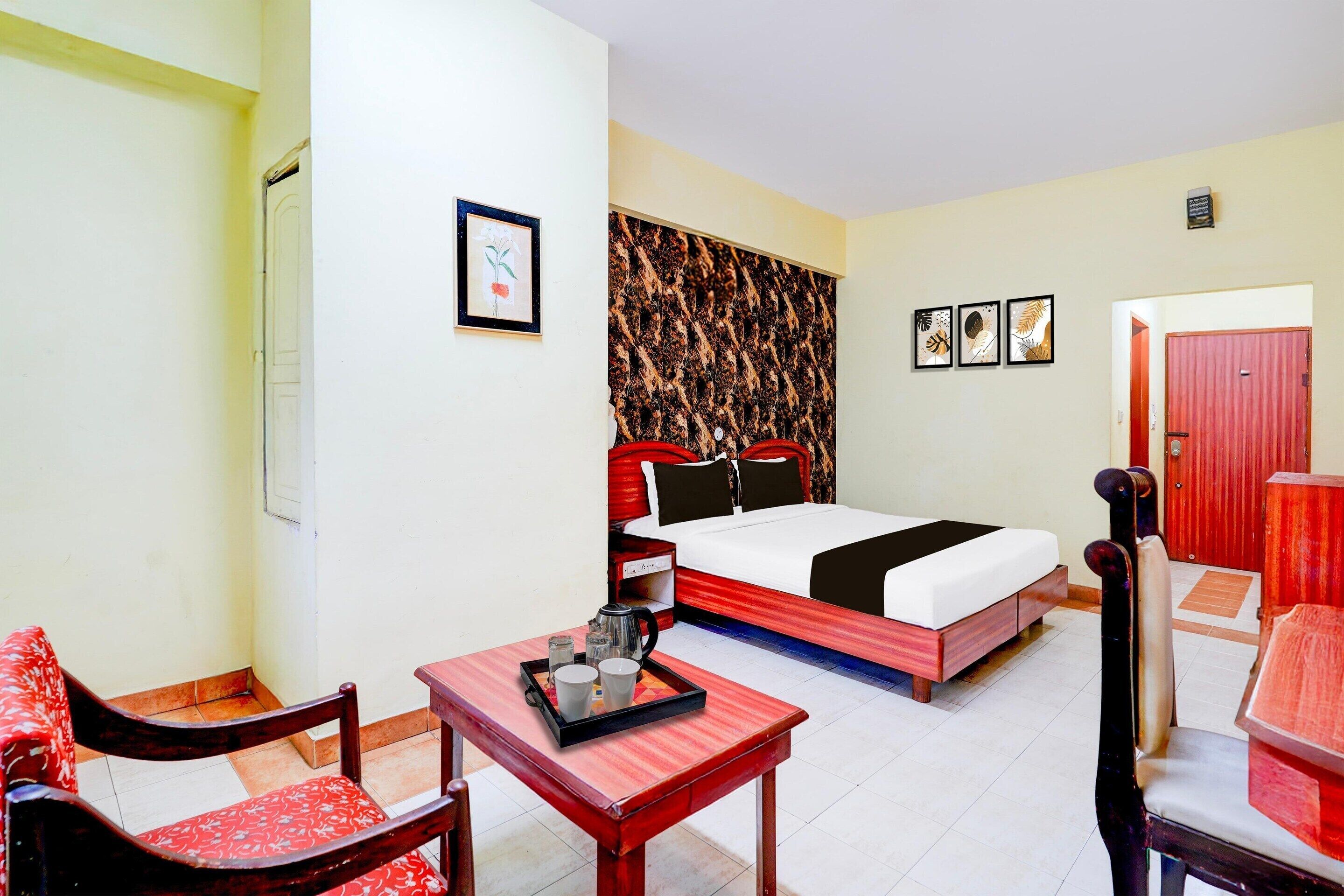 Quarto de hotel com cama, mesa com bandeja de objetos, uma cadeira e uma porta vermelha.