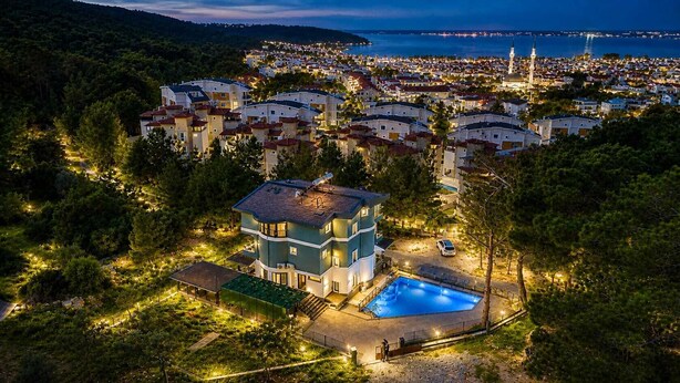 Um edifício de vários andares com piscina, rodeado por árvores e vista para uma cidade ao longe.