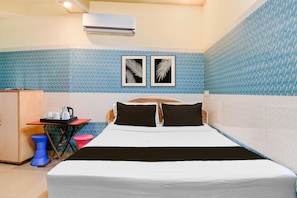 Quarto de hotel com uma cama, uma pequena mesa com um chaleira, um banquinho azul e um banquinho vermelho.