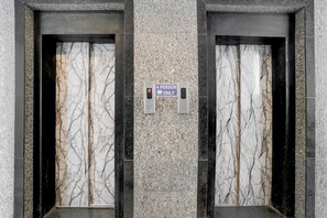 Dois elevadores com paredes de mármore e um aviso indicando '4 PESSOAS NO MÁXIMO'.