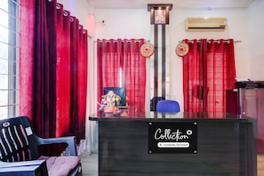 Balcão de recepção com placa indicando serviço de uma empresa, uma cadeira azul, uma cadeira roxa e cortinas vermelhas com padrão decorativo.