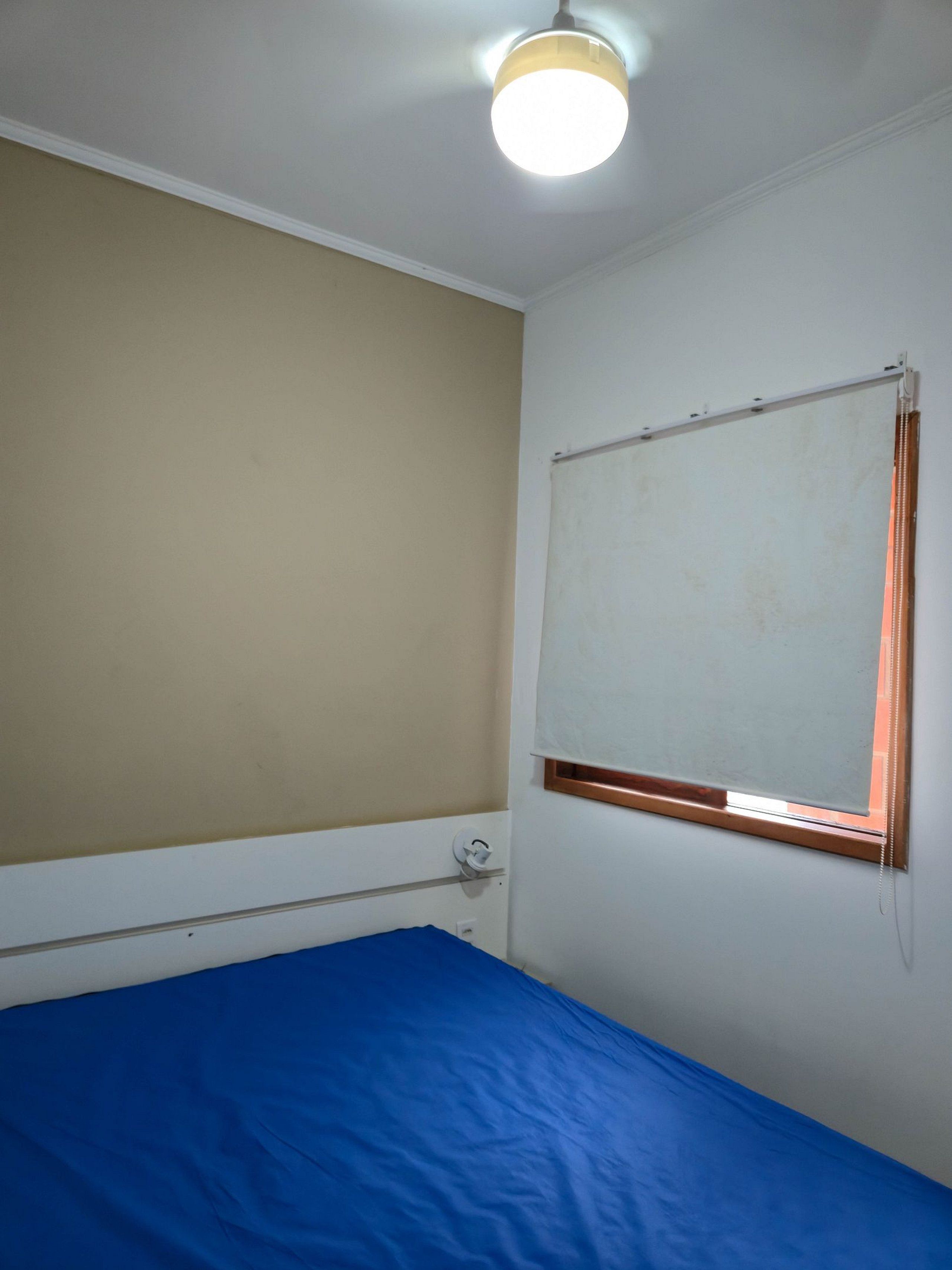Quarto com cama, luminária no teto, janela com persianas e radiador fixado na parede.