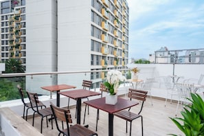 Terraço no telhado com mesas e cadeiras, um vaso de planta e vista para um edifício alto.