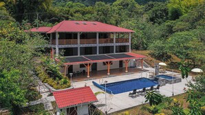 Um edifício de dois andares com telhado vermelho, piscina e área de varanda coberta.