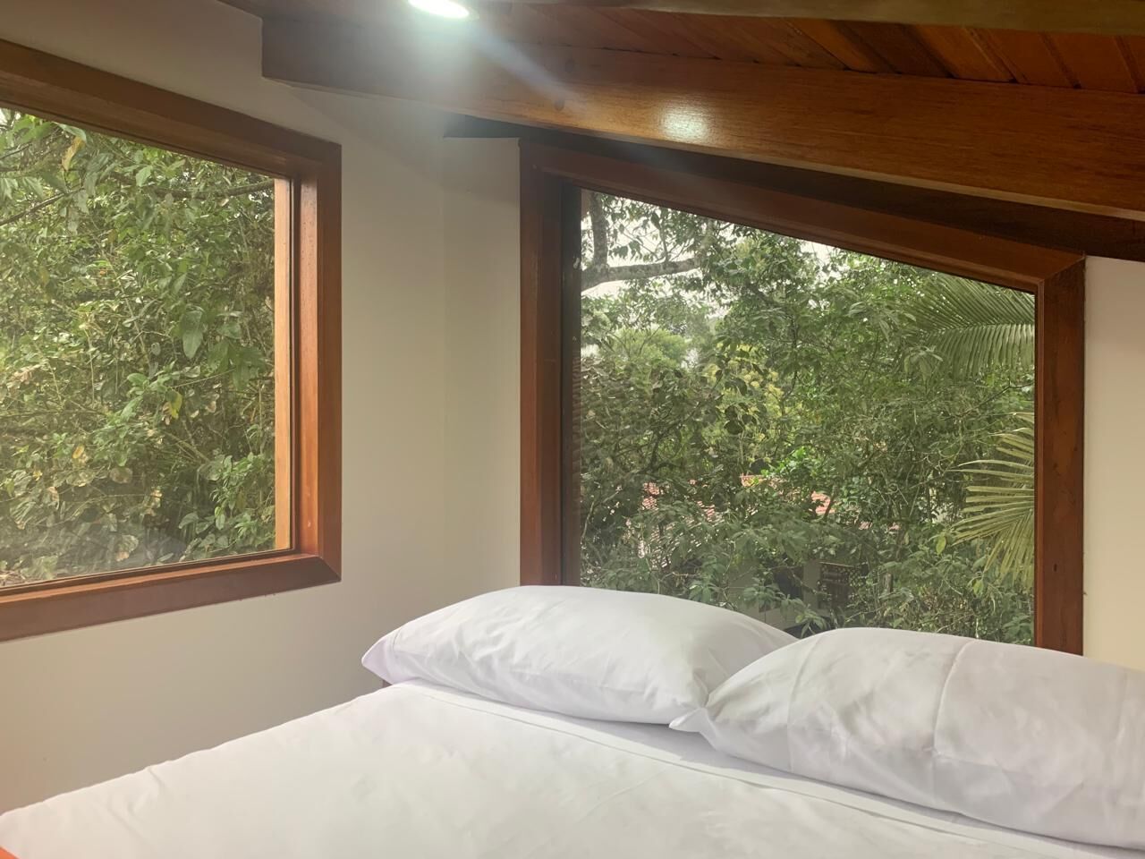 Quarto com cama, almofadas brancas e janela com vista para área verde.