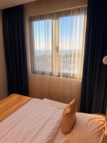Quarto de hotel com cama, janela com cortinas finas e vista para a paisagem urbana.