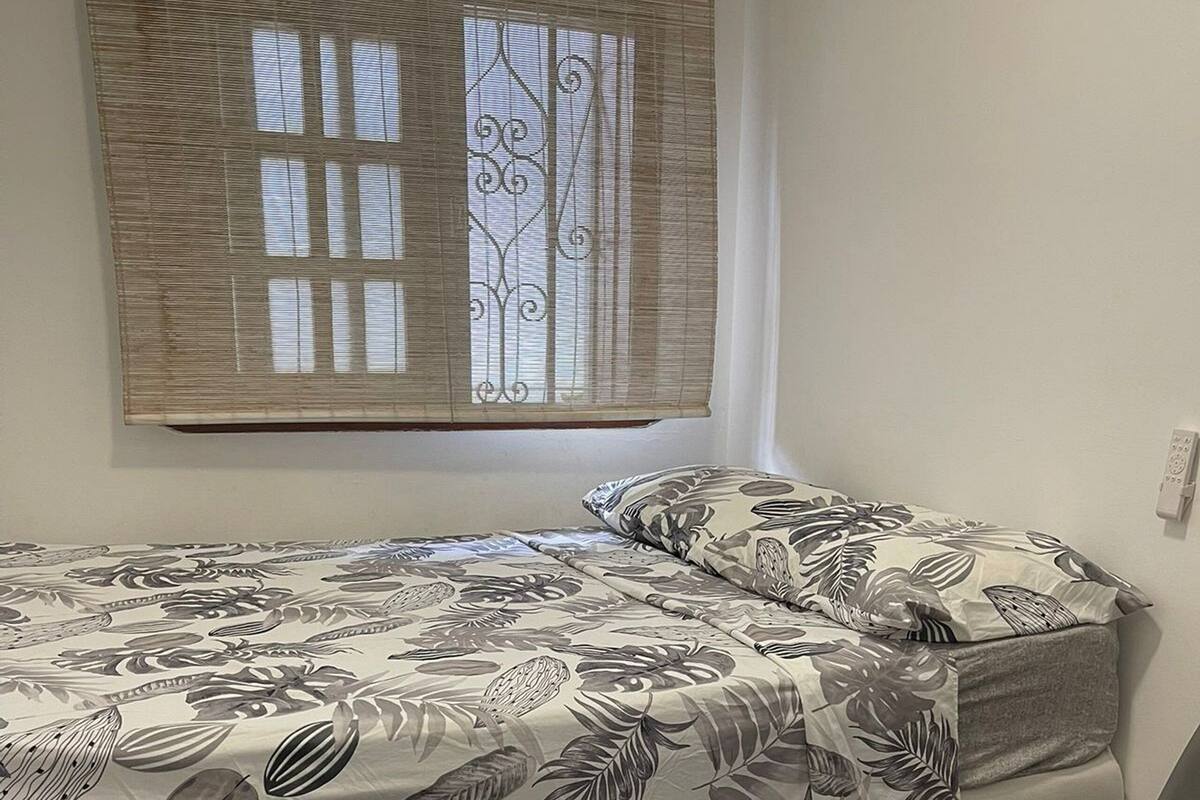 Uma cama de solteiro com colcha florida, uma janela com mosquiteiro e uma cadeira.
