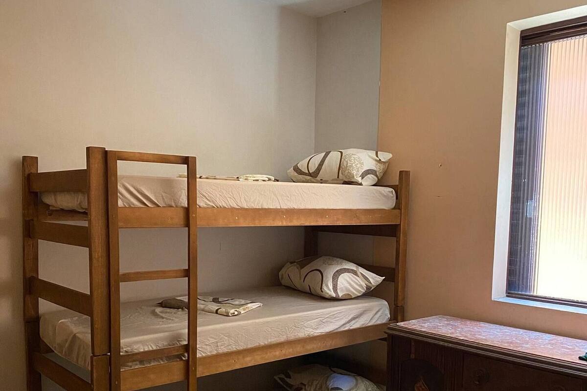 Uma cama beliche com escada de madeira, uma cômoda com gavetas e uma janela com persianas.