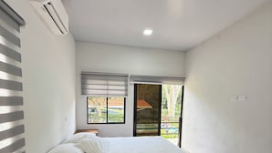 Quarto com cama, janela com persianas, ar-condicionado e vista para o exterior.