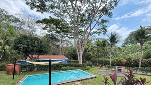 Piscina cercada por um gramado bem cuidado, uma pequena construção de tijolos e várias árvores.