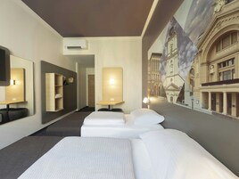 Quarto de hotel com duas camas, um espelho grande, ar condicionado fixado na parede e um mural de um edifício.