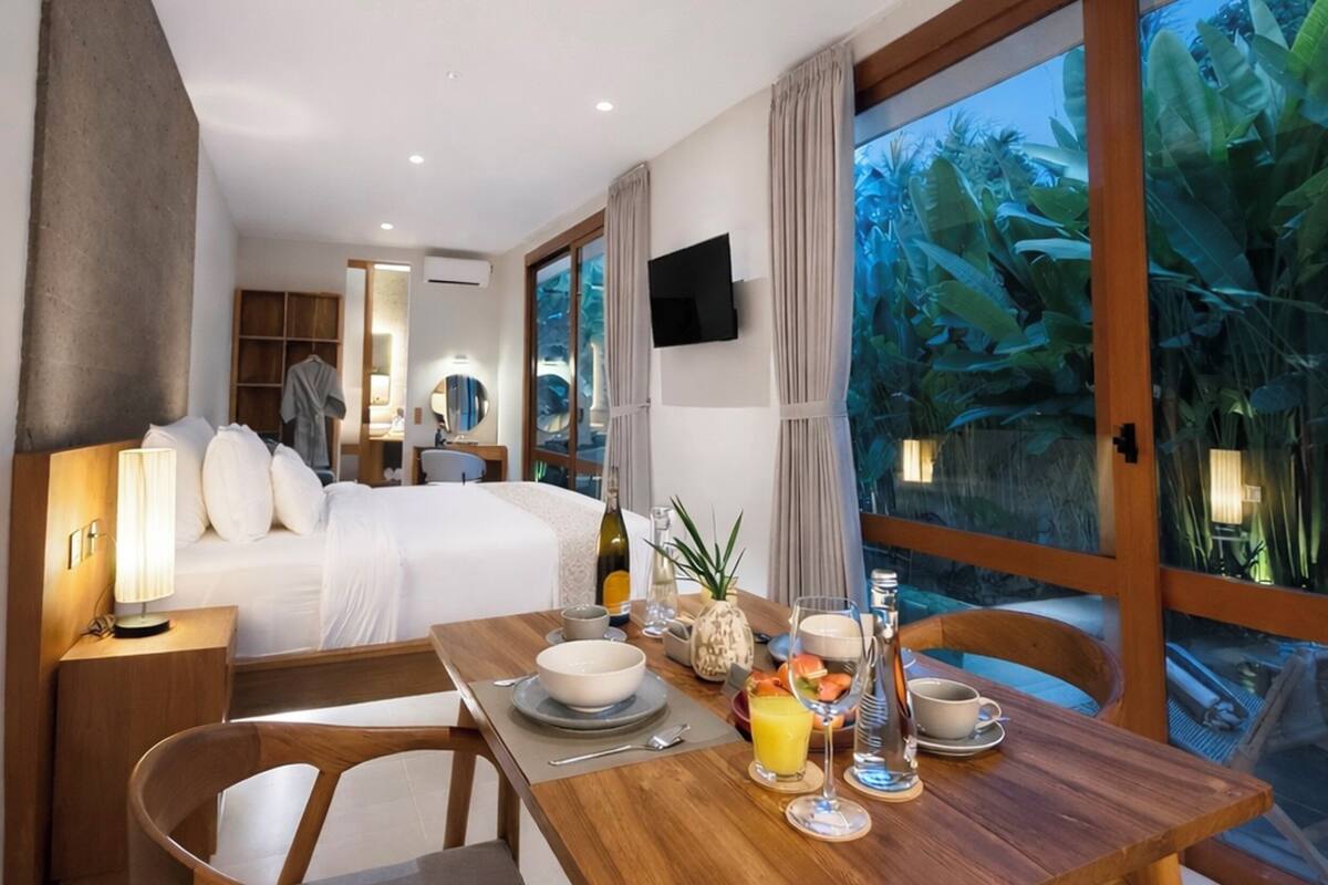 Quarto de hotel com cama, mesa de jantar com café da manhã, televisão e uma grande janela com vista para o verde.
