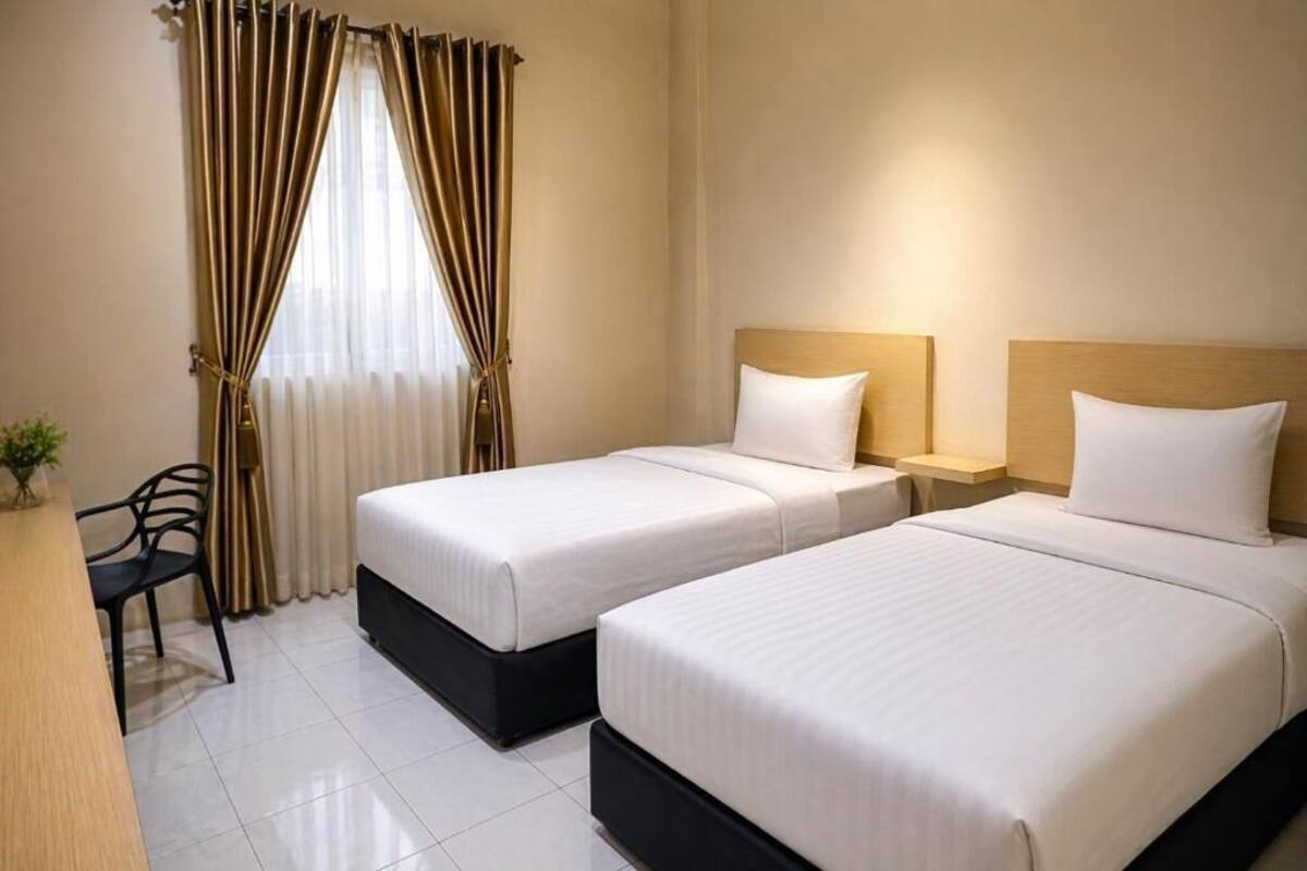 Quarto de hotel com duas camas, uma escrivaninha, uma cadeira e uma janela com cortinas.