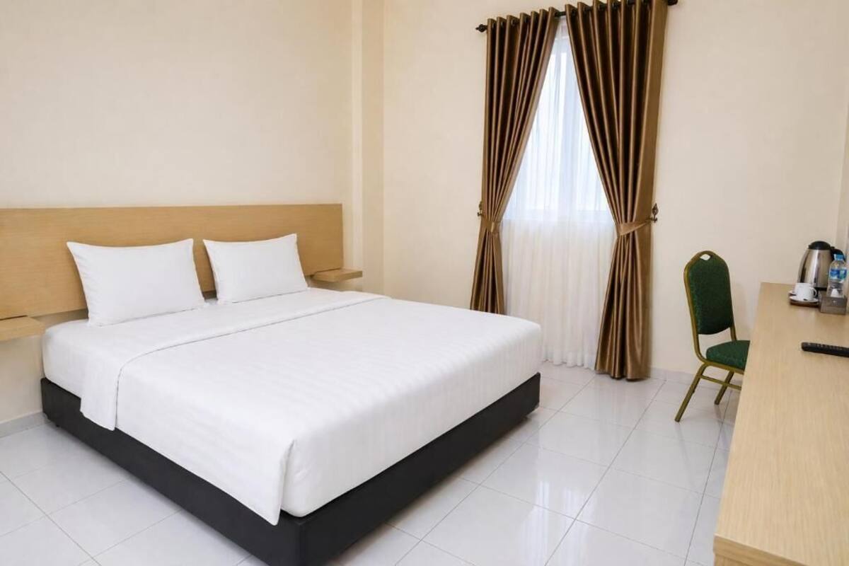 Quarto de hotel com cama, escrivaninha, cadeira e janela com cortinas.
