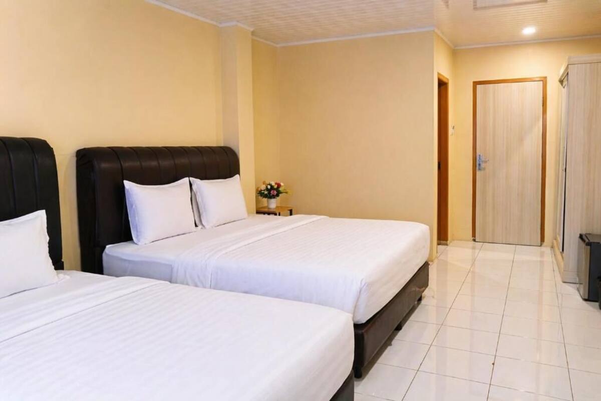 Quarto de hotel com duas camas, uma mesa de cabeceira com um vaso, um guarda-roupa e uma porta.