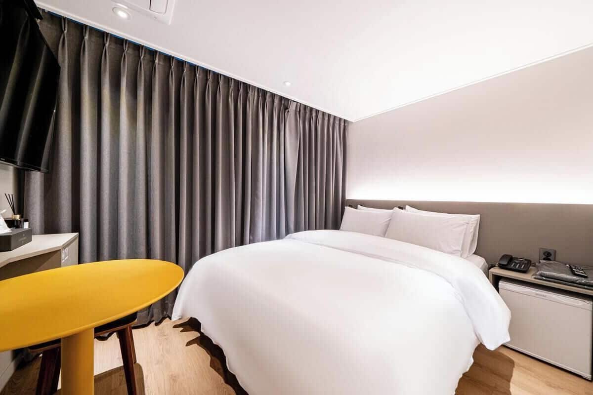 Quarto de hotel moderno com cama, mesa de cabeceira e pequena escrivaninha com cadeira.