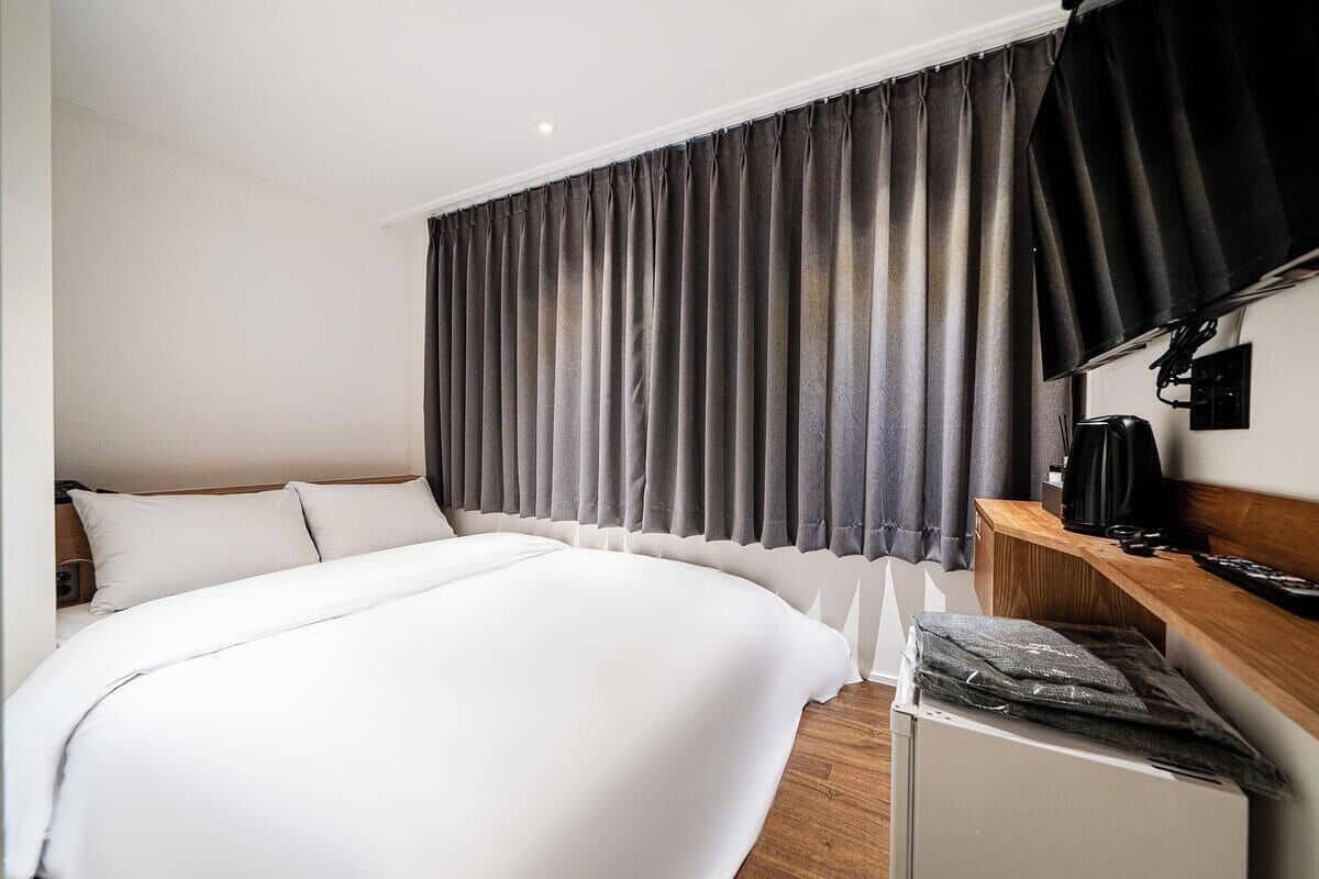 Quarto de hotel com uma cama, uma mesa de trabalho, uma máquina de café e uma televisão fixada na parede.