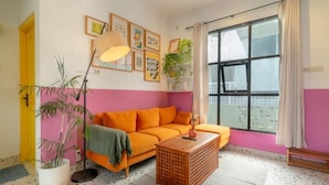 Quarto com esquema de cores rosa e amarelo, apresentando um sofá, uma mesa pequena, um abajur e uma janela com cortinas.