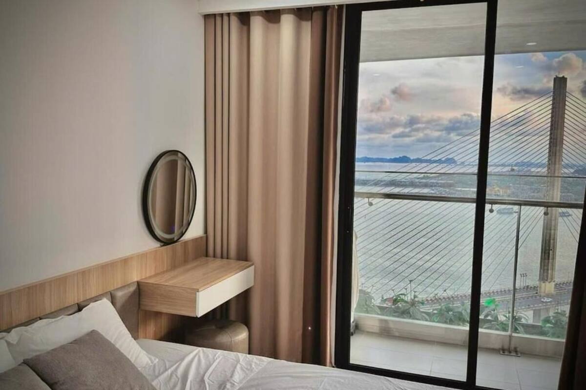 Quarto de hotel com cama, mesa de cabeceira, espelho e janela com vista para uma ponte e paisagem urbana.