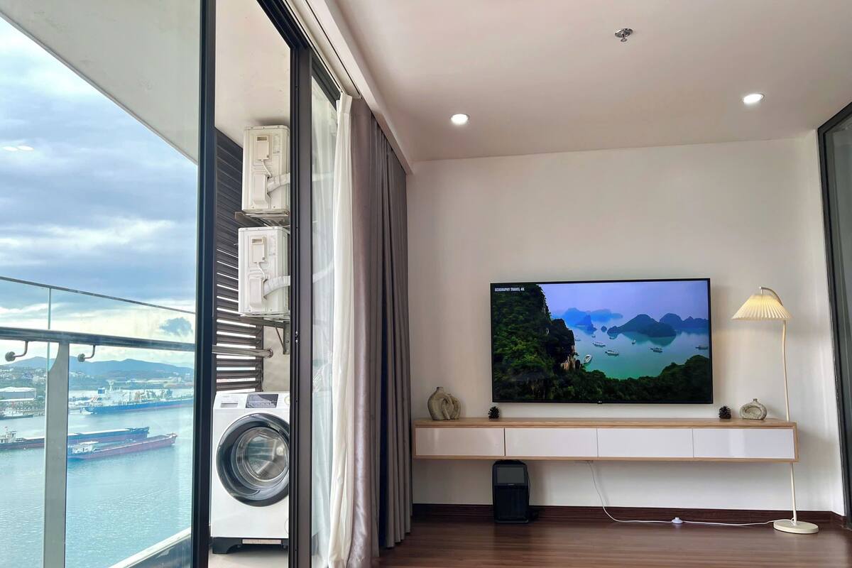 Quarto com uma janela ampla com vista para um corpo de água, máquina de lavar, televisão na parede e uma pequena mesa com abajur.