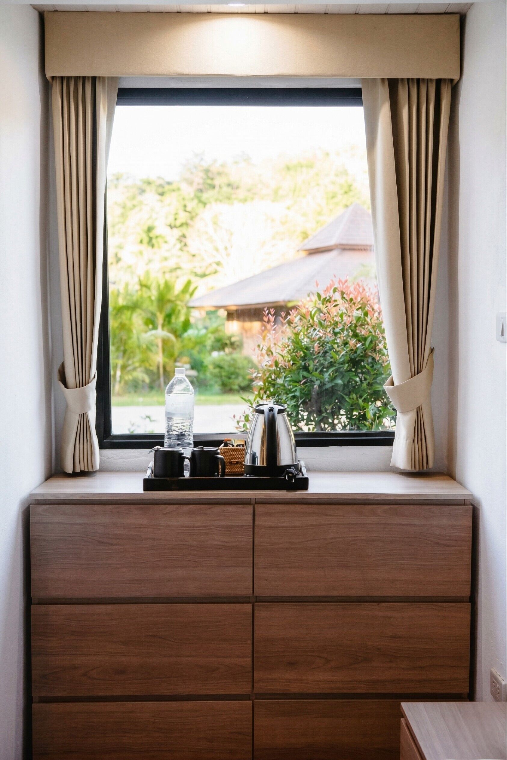 Uma janela com cortinas, uma garrafa de água, uma chaleira e um tabuleiro sobre uma cômoda de madeira.
