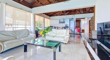Uma sala de estar ampla com uma mesa de centro de vidro, sofás brancos e uma cozinha ao fundo.