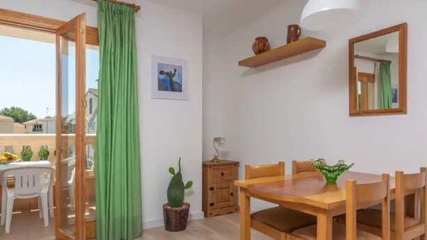 Uma área de refeições com mesa e cadeiras de madeira, cortina verde, planta de cacto, armário de madeira e um espelho na parede.