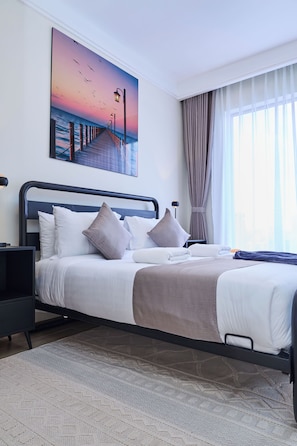 Quarto de hotel moderno com cama, mesa de cabeceira, pintura de um pôr do sol sobre um cais e janela com cortinas.