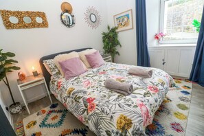 Quarto com colcha florida, abajures, um vaso de planta e janela com cortinas.