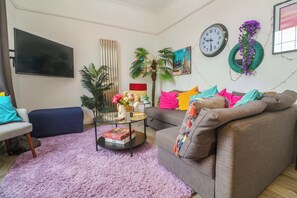 Uma sala de estar com sofá, mesa de centro, televisão e um relógio na parede.
