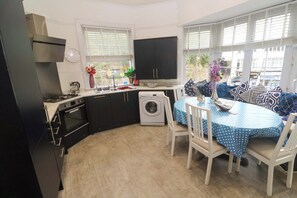 Cozinha compacta com fogão, forno e máquina de lavar roupa, área de refeições com mesa e cadeiras, e janelas amplas com persianas.