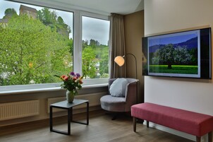 Um quarto com uma janela grande, uma cadeira, um banco, uma mesa com um vaso de flores e uma televisão na parede.