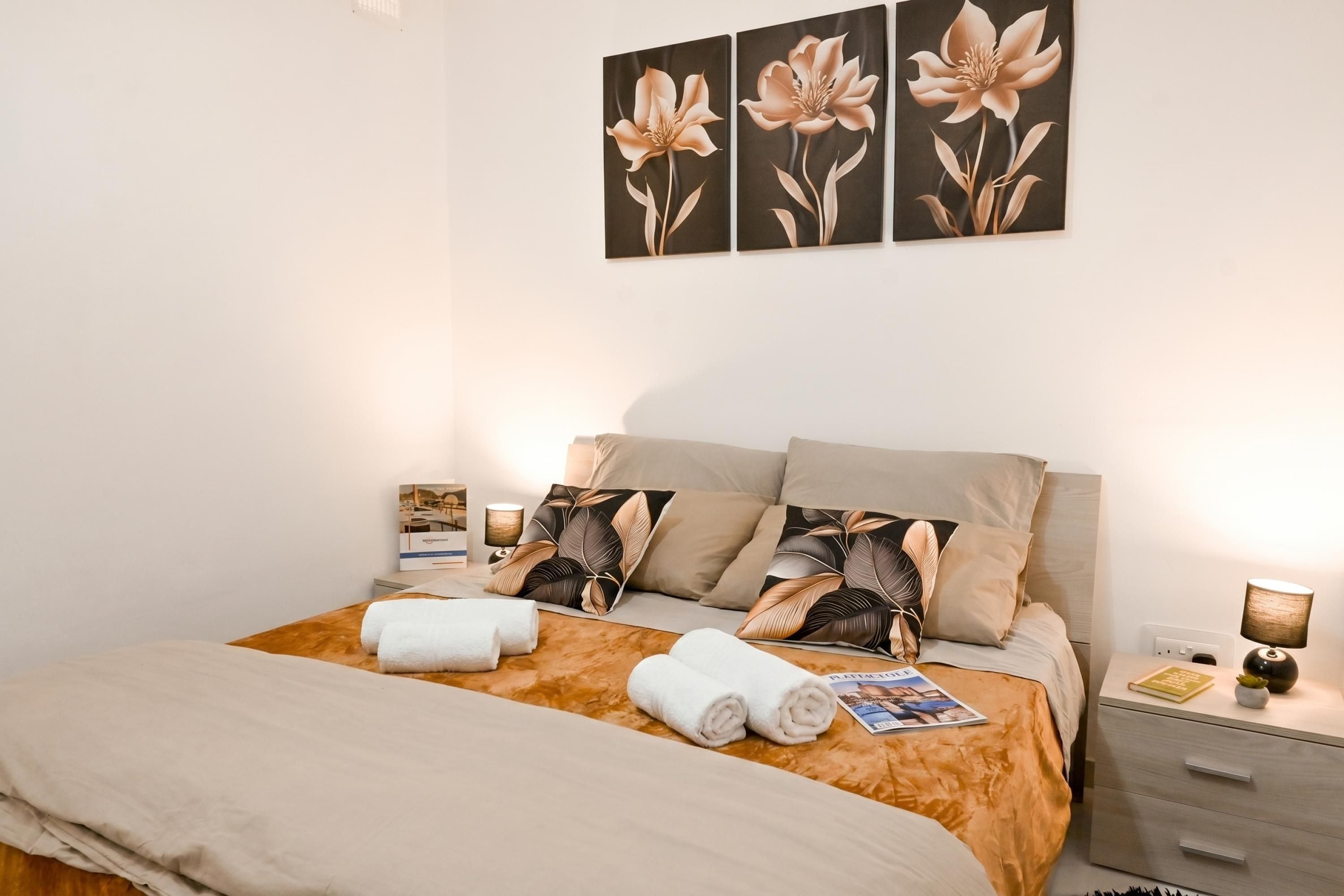 Cama bem arrumada com toalhas, mesa de cabeceira com abajur e parede com duas pinturas florais.