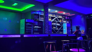 Uma área de bar com bancos, prateleiras cheias de garrafas e um atendente atrás do balcão.