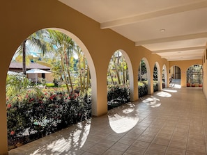 Um corredor com tetos abobadados, soalho em azulejos e vista para um jardim com diversas plantas e flores.