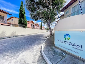 Um imóvel residencial com uma rua pavimentada, uma parede branca com uma placa e uma árvore.