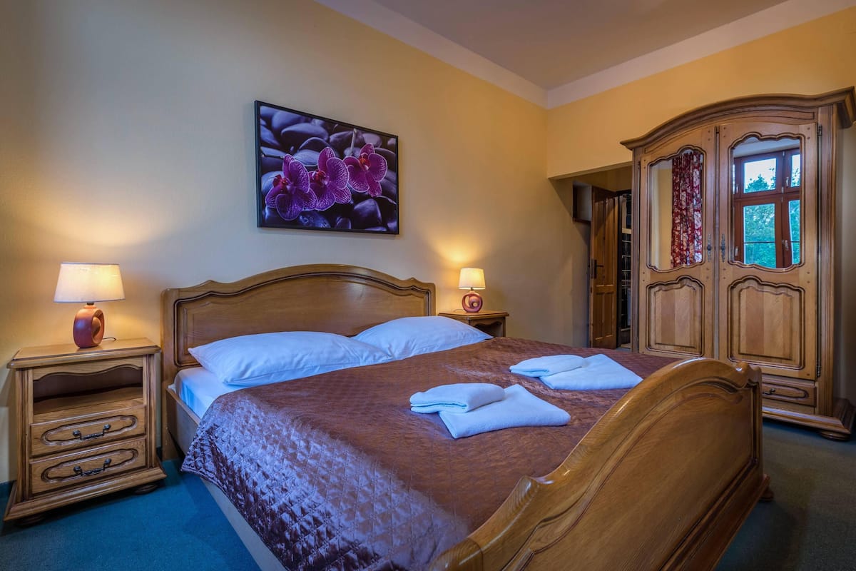 Cama bem arrumada com almofadas, mesa de cabeceira de madeira com abajur, guarda-roupa de madeira e um quadro na parede.