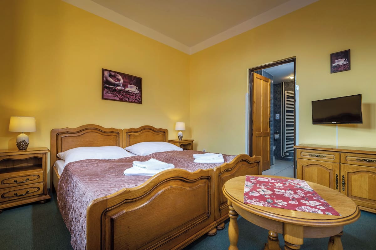 Quarto de hotel com cama de madeira, mesas de cabeceira com luminárias, televisão e porta para o banheiro.