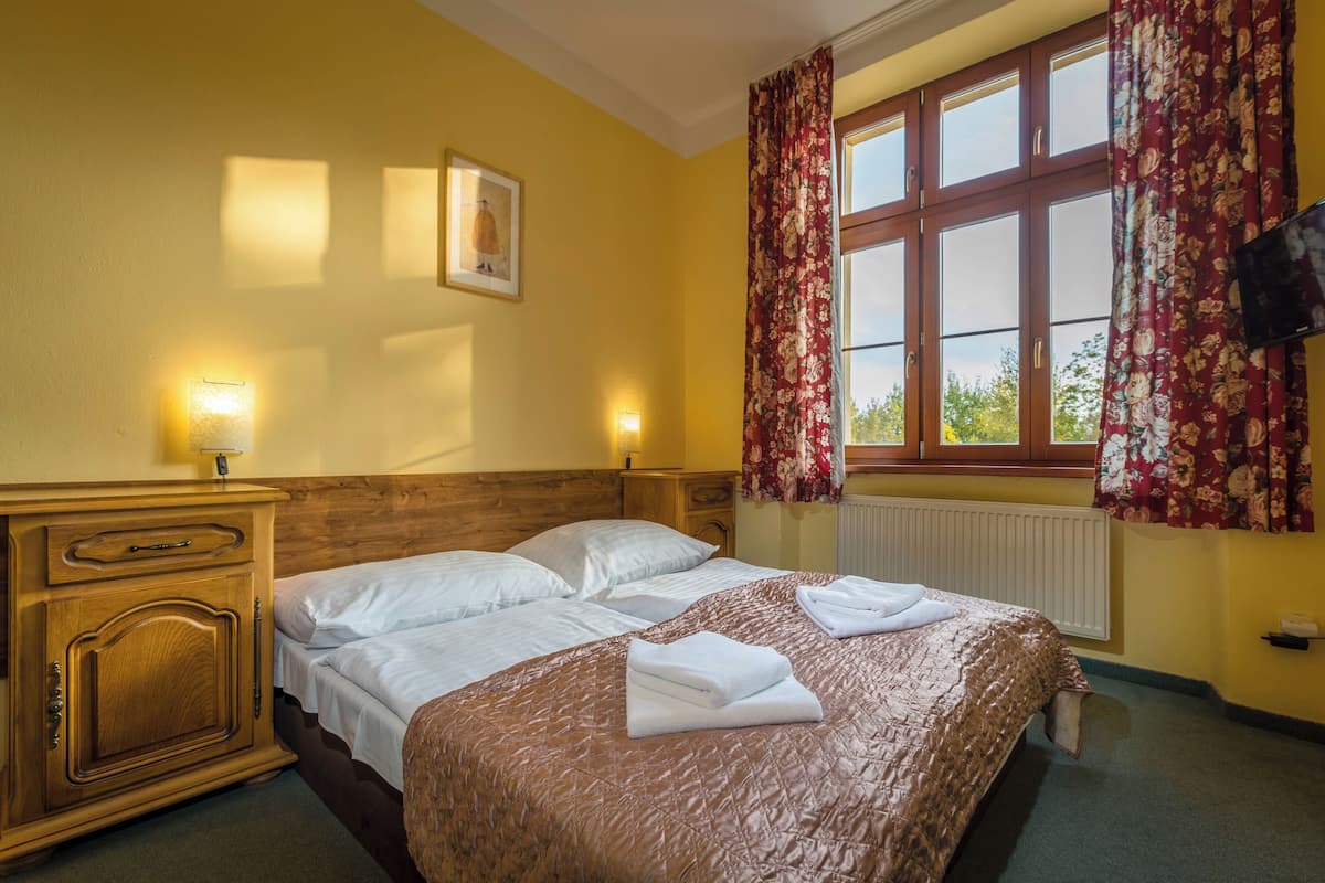 Quarto de hotel com cama de casal, cabeceira de madeira, duas mesas de cabeceira com luminárias, uma janela com cortinas e um radiador.