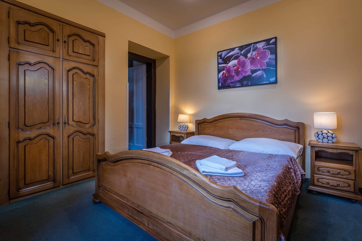 Um quarto com uma cama de madeira, duas mesas de cabeceira com abajures, uma porta e um quadro de flores pendurado na parede.