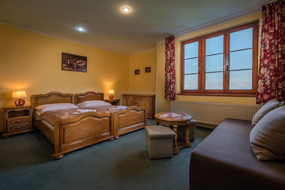 Quarto com cama de casal, mesas de cabeceira, mesa redonda, sofá, janela com cortinas e radiador.