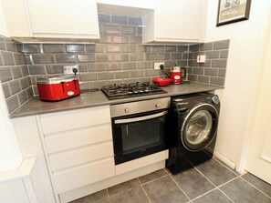 Uma cozinha compacta com fogão a gás, uma torradeira vermelha, máquina de lavar e painel de azulejos cinzas.