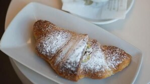 Um croissant sobre um prato branco, uma xícara de café com arte de latte e uma mão alcançando o café.